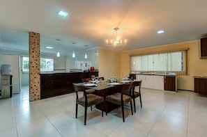 Uma ampla sala de jantar com uma mesa grande posta para seis pessoas, rodeada por cadeiras. Uma cozinha é visível ao fundo, com eletrodomésticos modernos e uma grande janela que oferece luz natural.