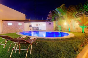 Uma piscina com espreguiçadeiras, um terraço e um jardim bem iluminado com palmeiras.
