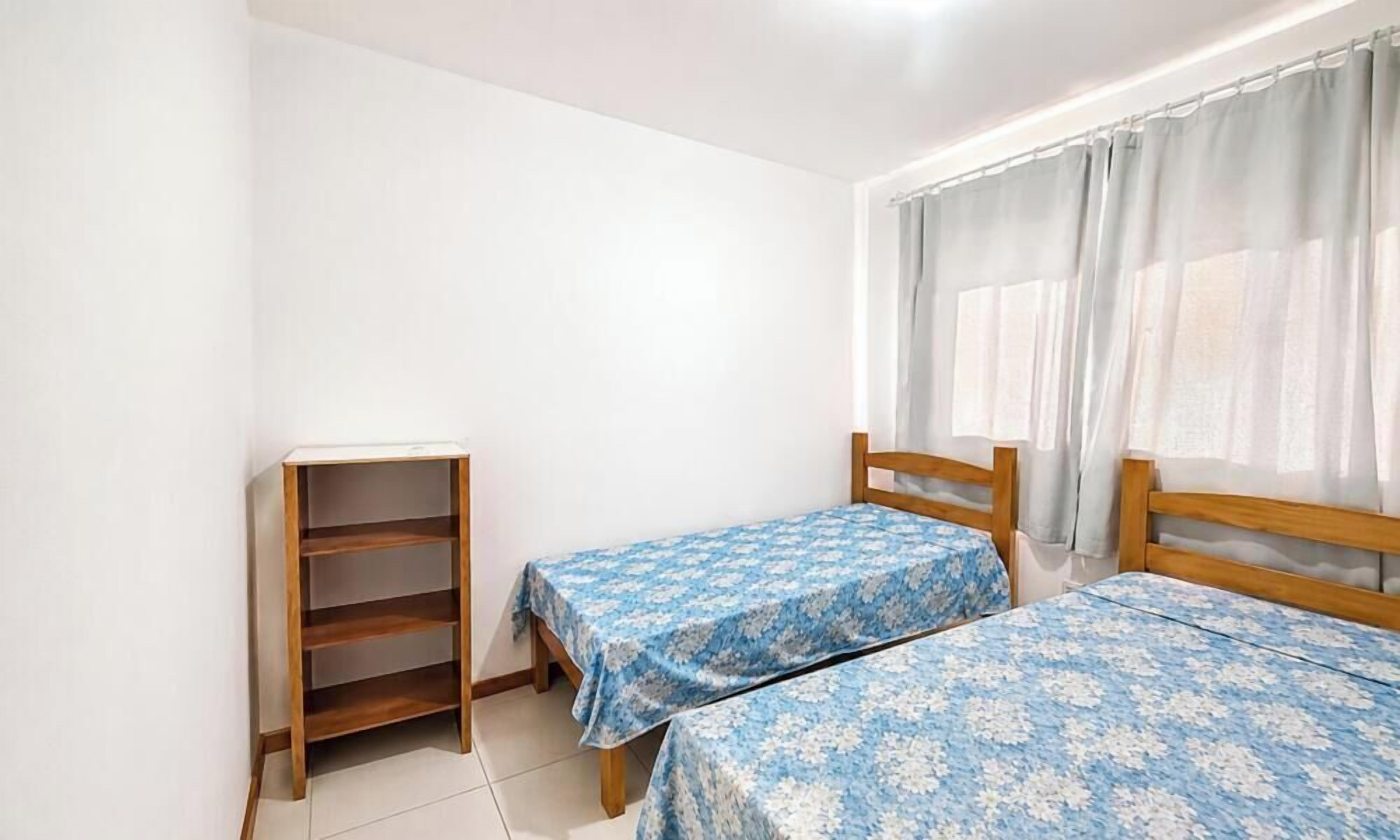 Quarto pequeno com duas camas de solteiro, uma estante de madeira e uma janela com cortinas leves.