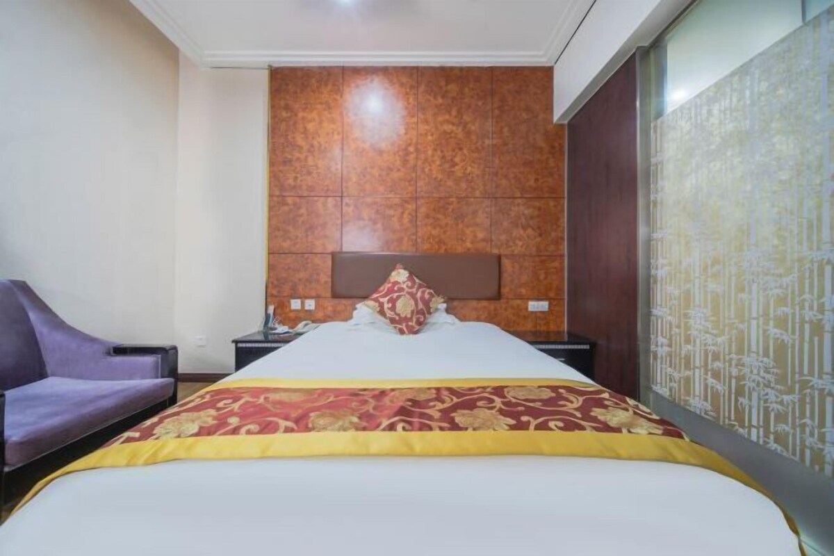 Quarto de hotel com uma cama, mesa de cabeceira, abajur, poltrona roxa e um painel decorativo na parede.