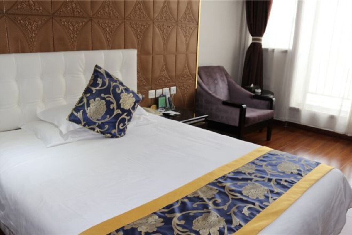 Quarto de hotel com uma cama grande, cabeceira com padrão geométrico, poltrona roxa e uma mesinha de cabeceira com abajur.