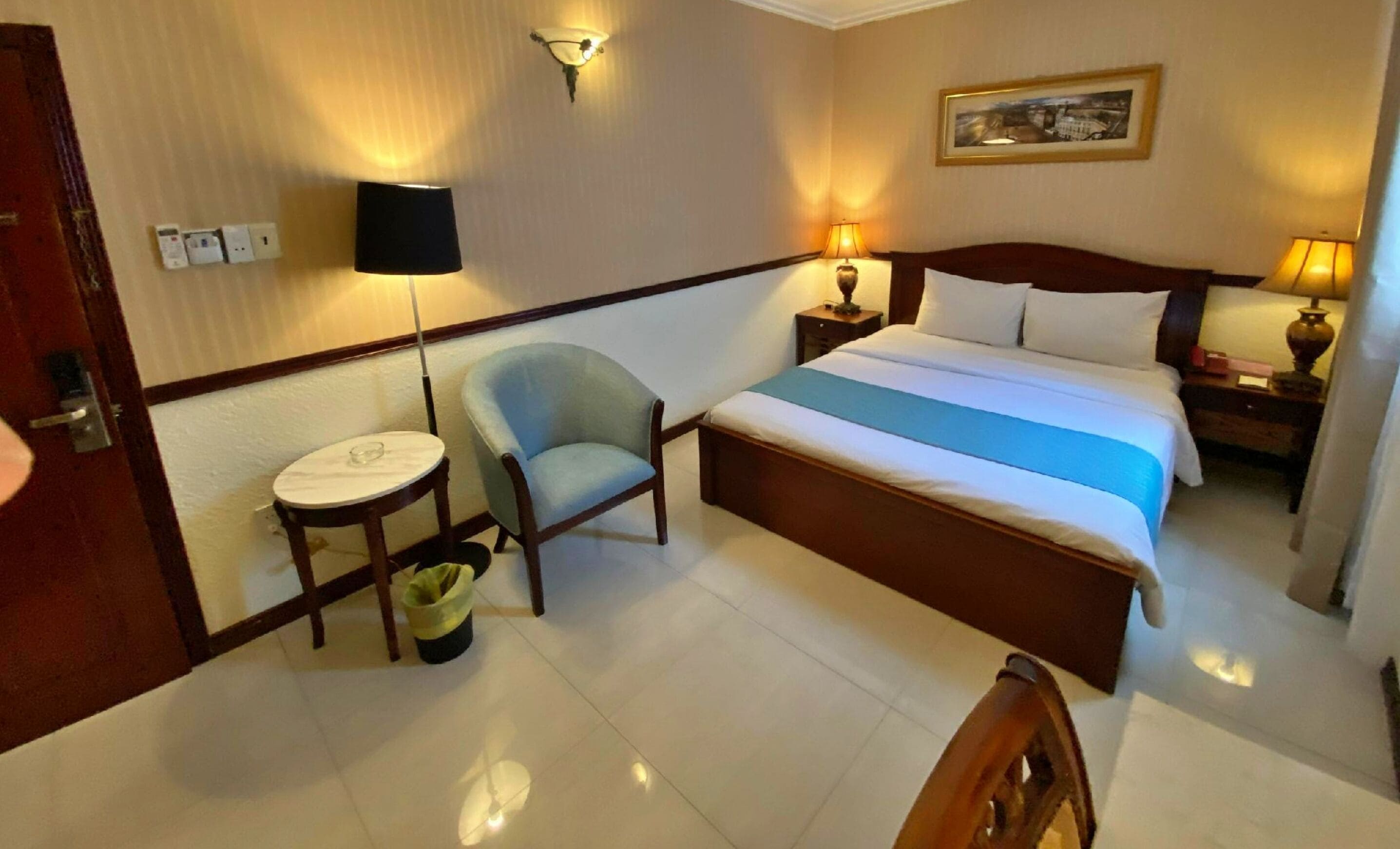 Quarto de hotel com cama, mesas de cabeceira, uma cadeira, uma mesinha e um cesto de lixo.