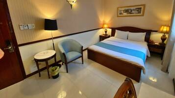 Quarto de hotel com cama, mesas de cabeceira, uma cadeira, uma mesinha e um cesto de lixo.
