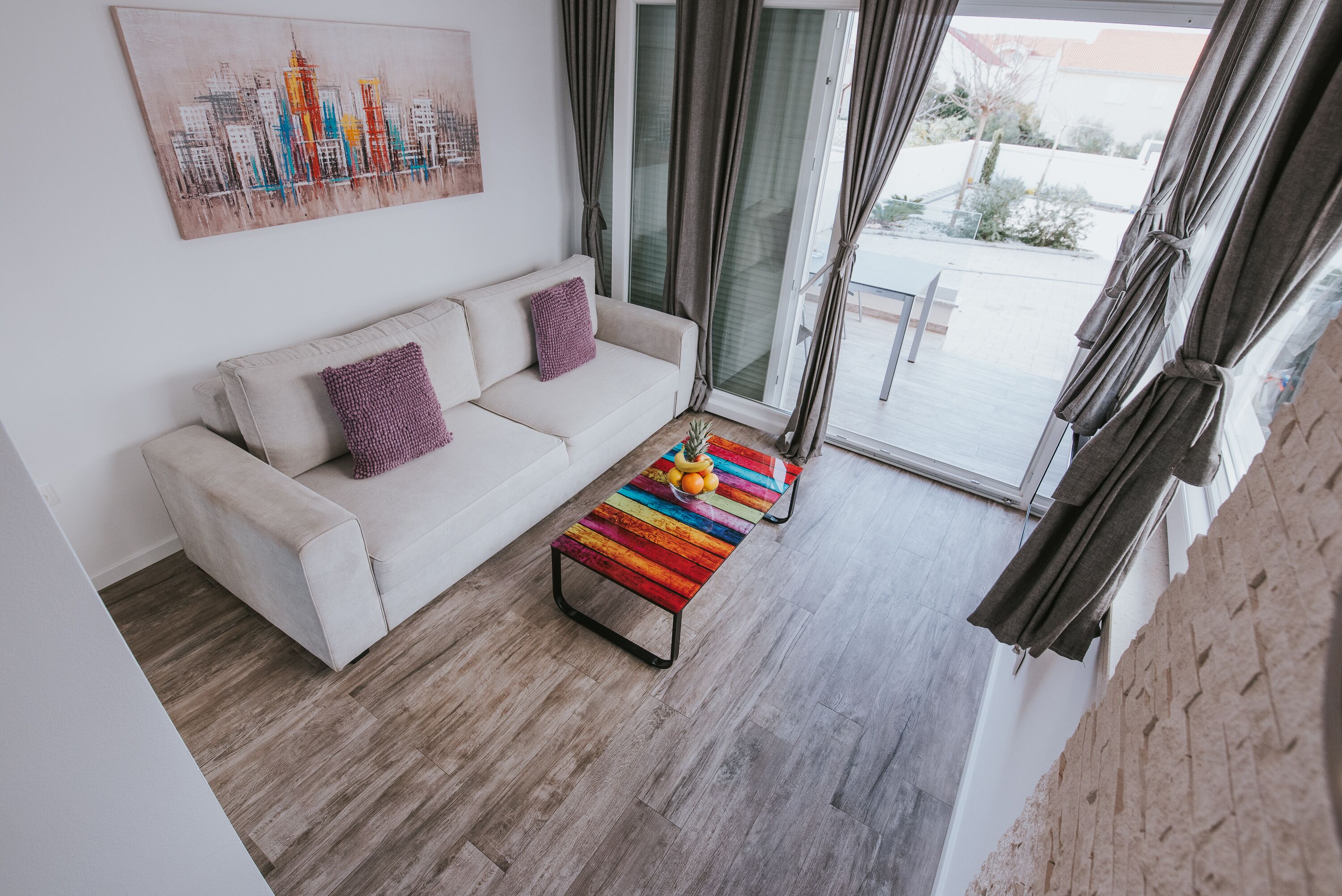 Uma sala de estar moderna com um sofá branco, mesa de centro listrada e colorida, e uma pintura com vista para a cidade na parede.