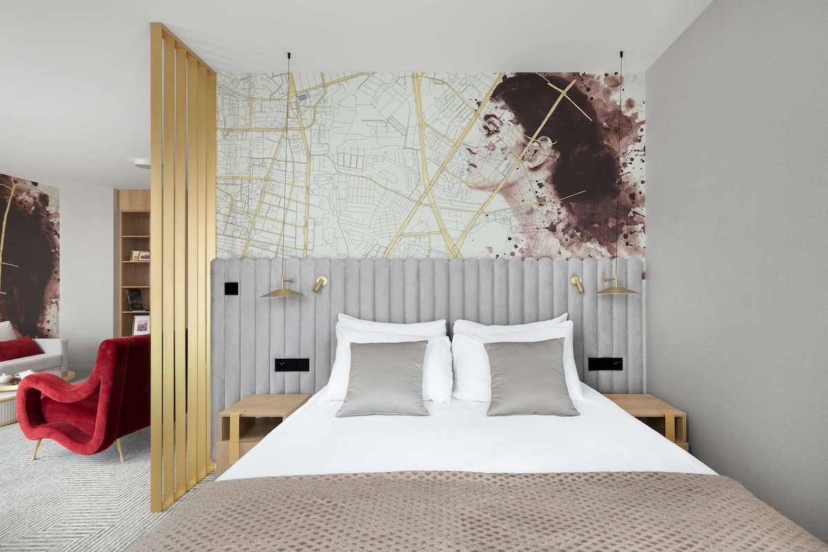 Quarto de hotel moderno com uma cama grande, um mapa na parede, uma cadeira vermelha e uma mesinha de madeira.
