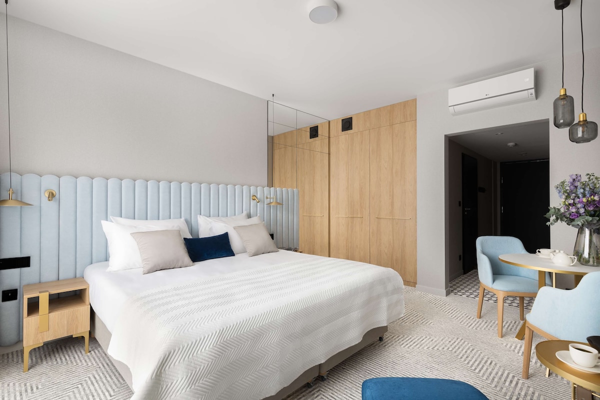Quarto de hotel moderno com uma cama grande, um guarda-roupa de madeira, uma pequena mesa com uma chávena e uma cadeira.