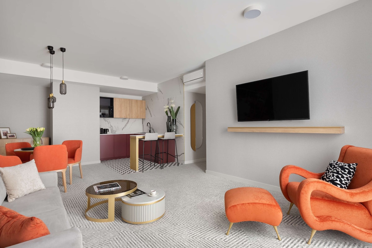 Uma sala de estar moderna com sofá, poltrona, banqueta e mesa de centro, além de uma TV fixada na parede.