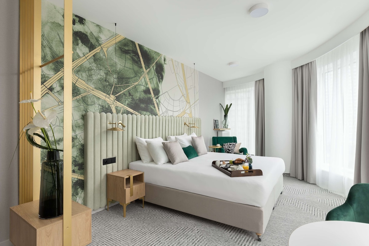 Quarto de hotel moderno com uma cama grande, uma mesinha de cabeceira, uma cadeira verde e uma janela ampla com cortinas.