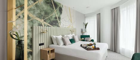 Quarto de hotel moderno com uma cama grande, uma mesinha de cabeceira, uma cadeira verde e uma janela ampla com cortinas.