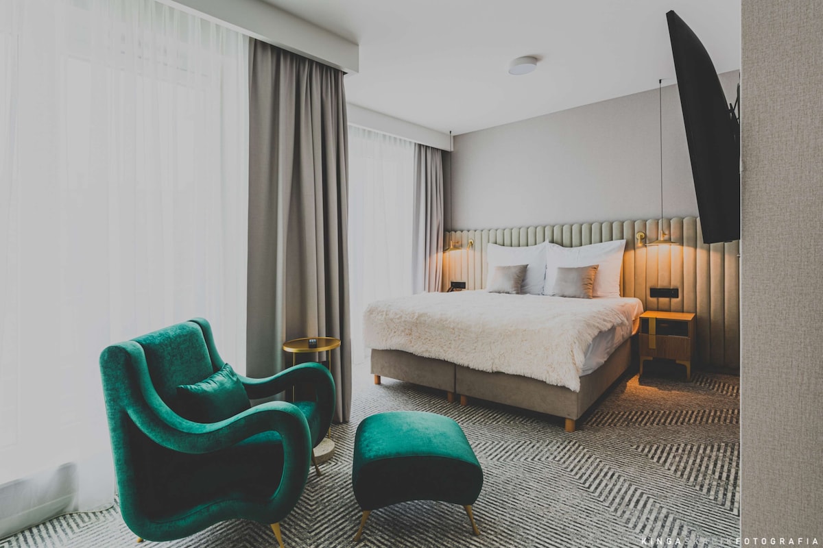 Quarto de hotel com cama, uma poltrona verde, uma mesa pequena e televisão fixada na parede.
