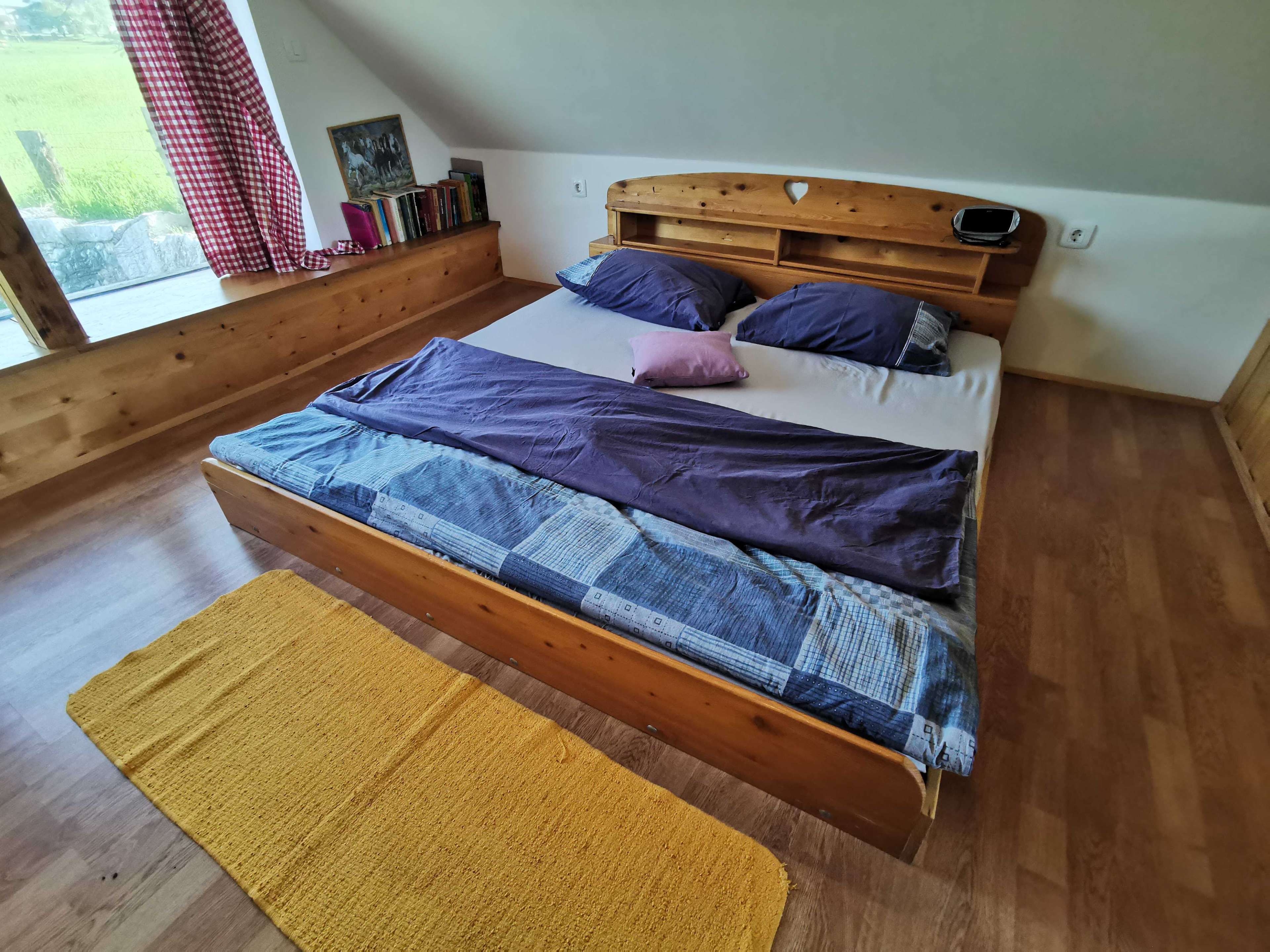 Quarto com cama de madeira, mesa de cabeceira, janela com vista e cortina xadrez vermelha e branca.