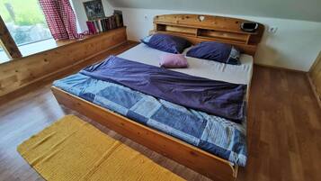 Quarto com cama de madeira, mesa de cabeceira, janela com vista e cortina xadrez vermelha e branca.