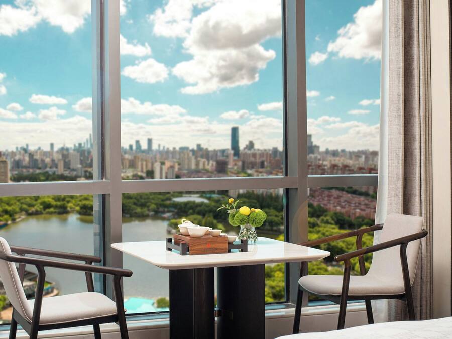 Quarto com uma janela ampla que oferece vista para a cidade e um rio. Há uma mesa com um vaso de flores e uma caixa de madeira. Duas cadeiras estão posicionadas ao redor da mesa.