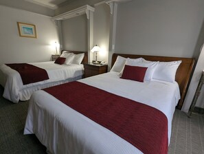 Duas camas de casal com lençóis brancos e mantas vermelhas, uma mesa de cabeceira com abajur, e um quadro na parede.