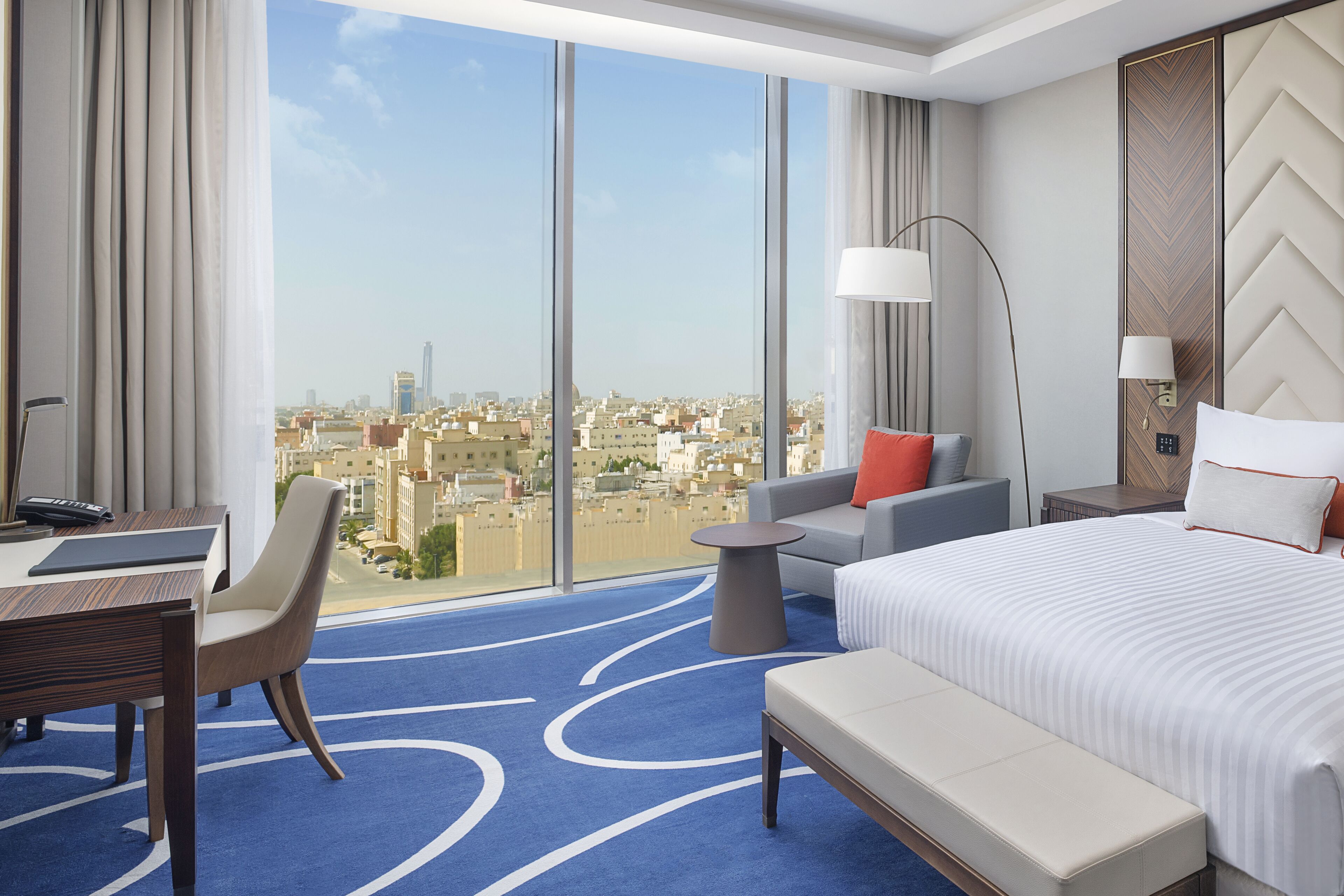 Quarto de hotel com uma janela grande, uma cama, uma escrivaninha e um espaço para sentar.