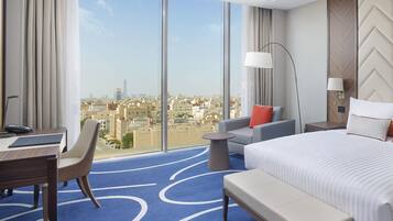 Quarto de hotel com uma janela grande, uma cama, uma escrivaninha e um espaço para sentar.