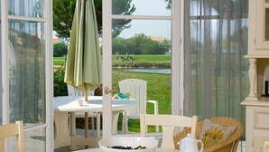 Quarto com uma mesa de jantar, cadeiras, uma cadeira de vime, uma tigela de frutas e vista para uma área gramada com árvores e um lago.