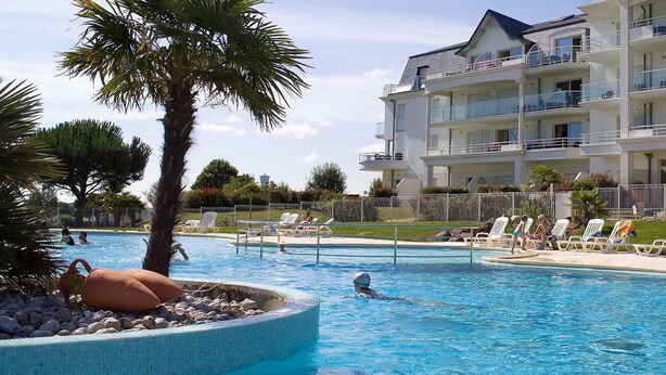Área de piscina com espreguiçadeiras, uma palmeira e um edifício com várias andares e varandas.