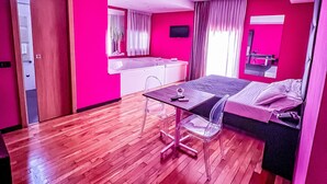 Quarto moderno com piso de madeira, cama, mesa pequena com cadeiras, televisão e banheiro com espelho e pia.