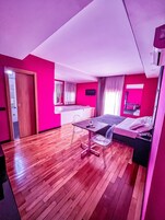 Quarto moderno com piso de madeira, cama, mesa pequena com cadeiras, televisão e banheiro com espelho e pia.