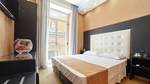 Quarto de hotel com uma cama grande, uma TV em um suporte preto, uma mesa de cabeceira e vista para um edifício através de uma porta de vidro.