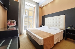 Quarto de hotel com uma cama grande, uma TV em um suporte preto, uma mesa de cabeceira e vista para um edifício através de uma porta de vidro.