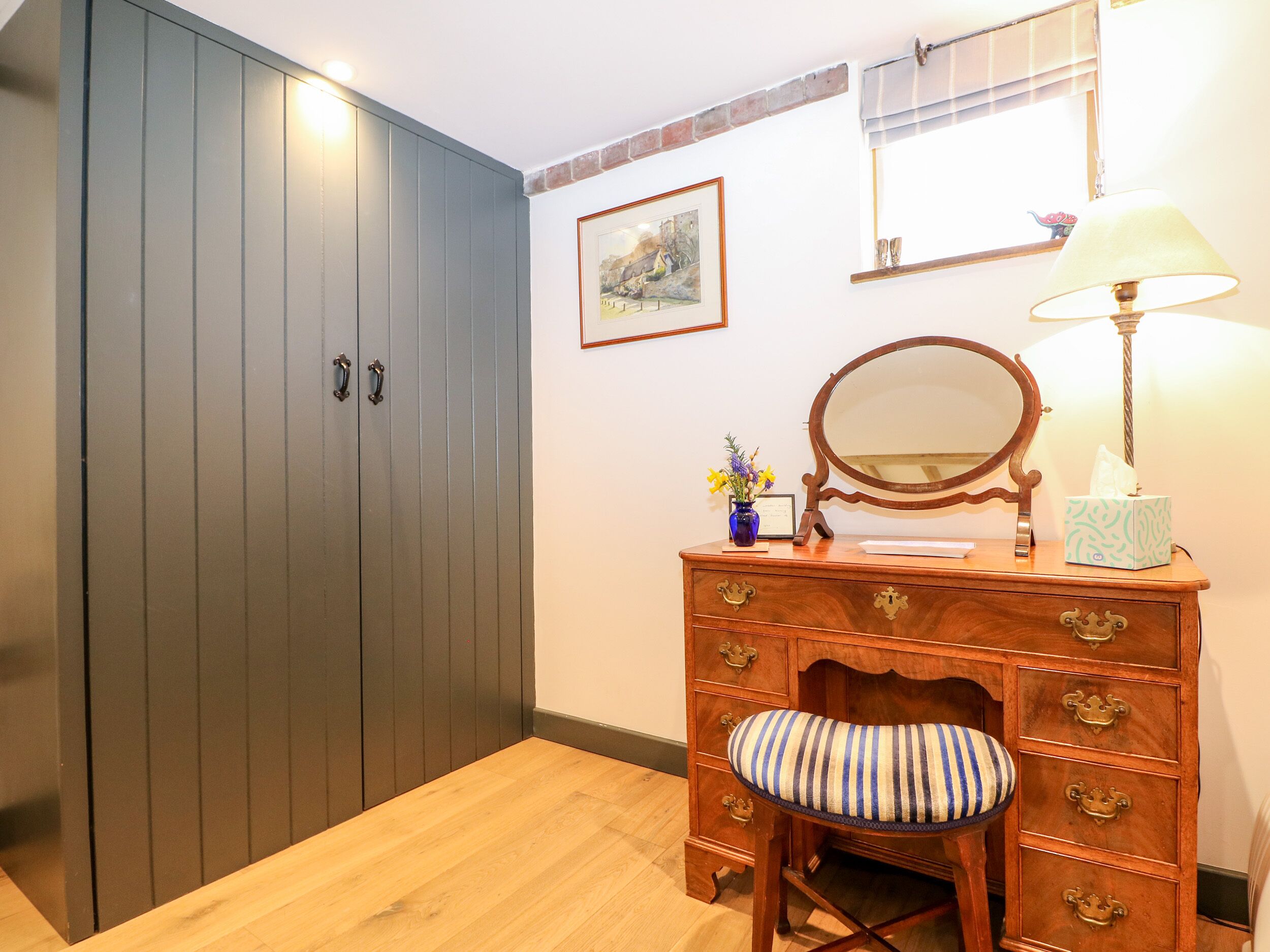 Quarto com penteadeira de madeira, espelho, luminária e quadro na parede.