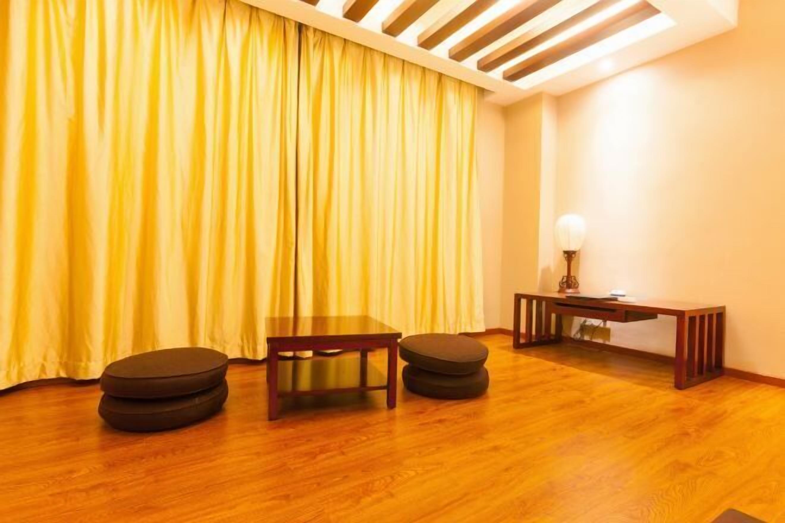 Quarto com piso de madeira, uma mesa, duas cadeiras, uma escrivaninha com abajur e cortinas amarelas.