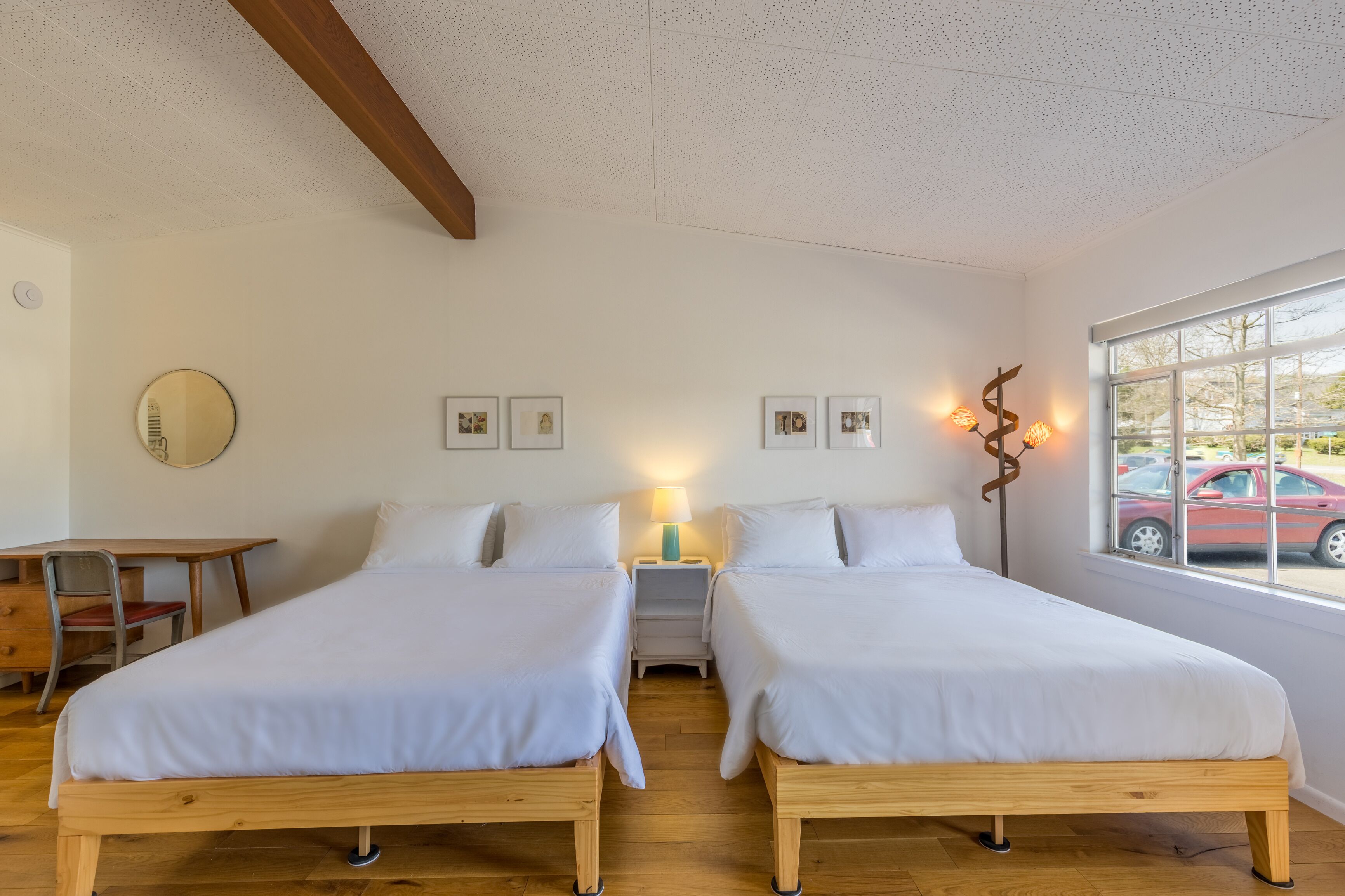 Quarto com duas camas de madeira, uma escrivaninha e uma cadeira. Há quadros nas paredes, um abajur e uma janela com vista para carros lá fora.