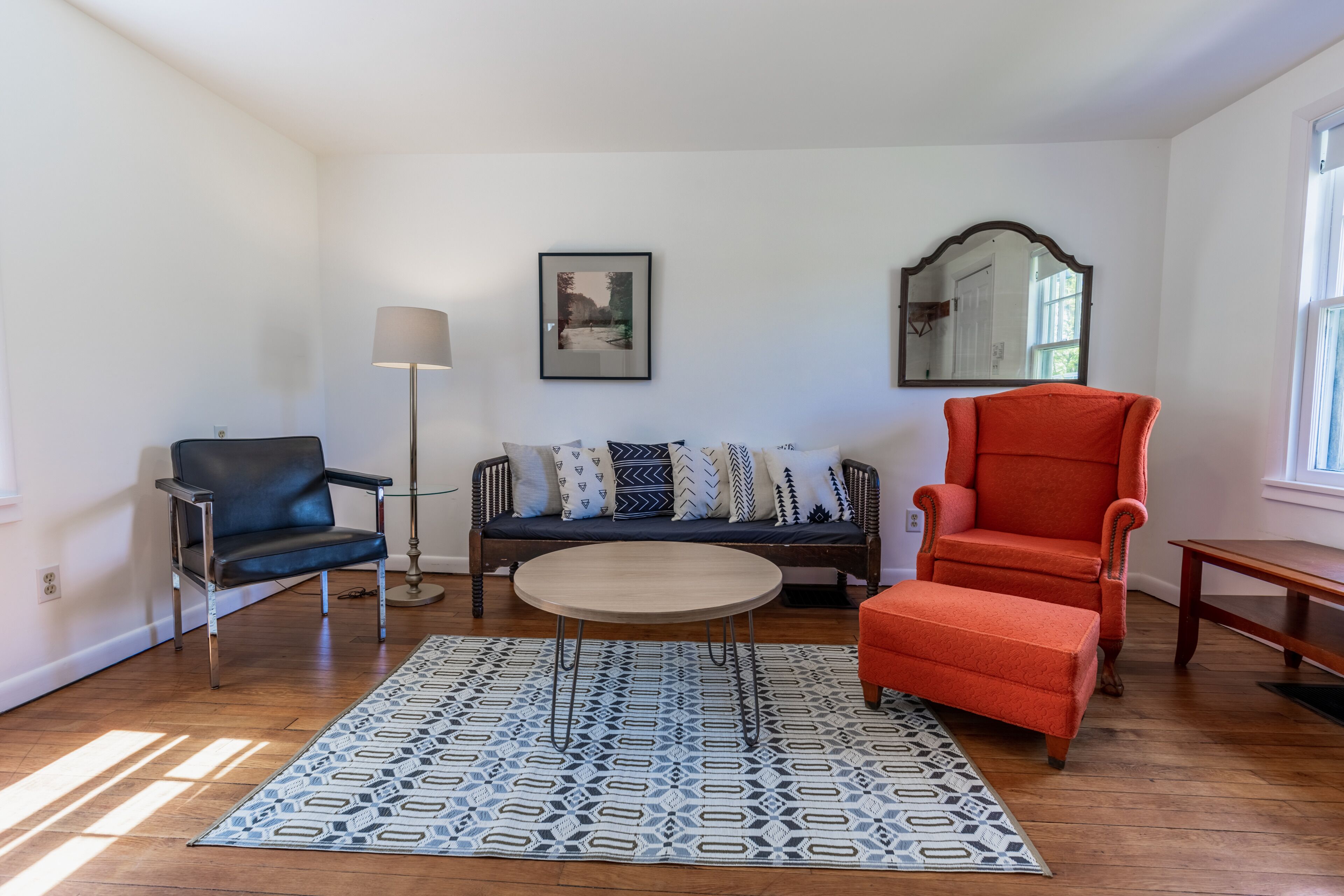 Uma sala de estar com sofá, duas cadeiras, mesa de centro, tapete estampado, luminária de chão e quadro na parede.