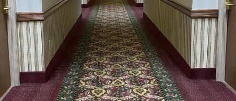 Um corredor longo e estreito com carpete estampado, paredes brancas e uma série de portas em ambos os lados.
