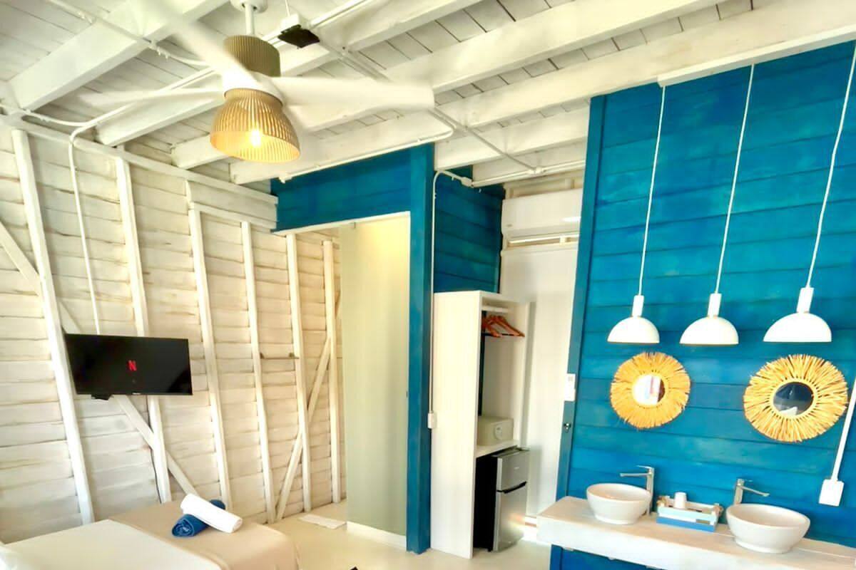 Quarto de hotel moderno com duas camas, televisão, banheiro com parede azul e uma pequena área de estar com mesa e cadeiras.