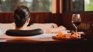 Uma pessoa relaxando em uma cadeira com um travesseiro de pescoço, uma taça de vinho tinto e uma tábua de madeira com comidas.