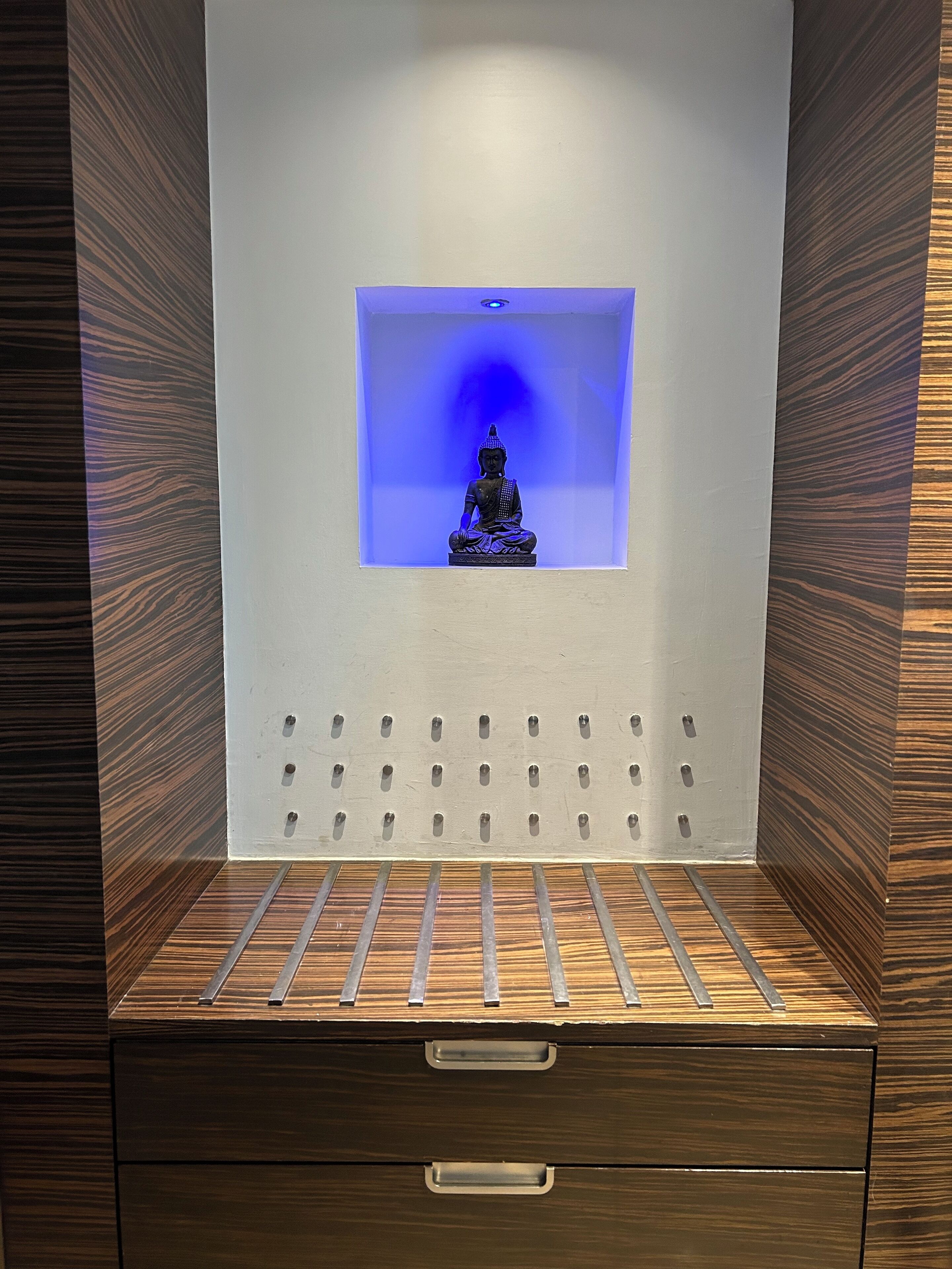 Estátua de Buda em um nicho iluminado por luz azul, cercada por uma grade de metal e uma gaveta de madeira abaixo.