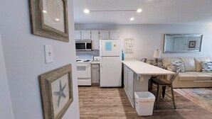Uma cozinha compacta com armários brancos, micro-ondas e geladeira. Há uma área de refeições com mesa e cadeiras, e um sofá com almofadas estampadas.
