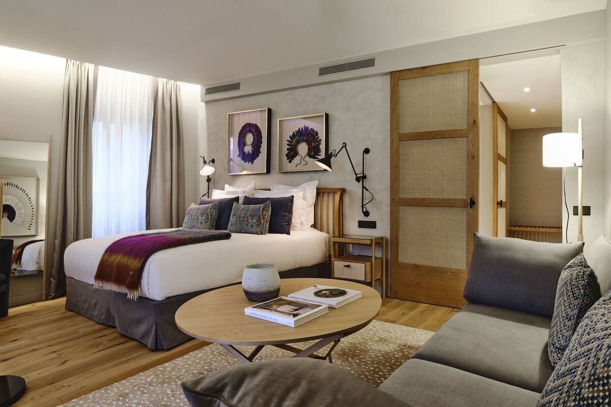 Quarto de hotel moderno com uma cama grande, um sofá, uma mesa de centro redonda e uma mesinha de cabeceira pequena. Há duas molduras com quadros na parede, um abajur e uma porta de madeira.