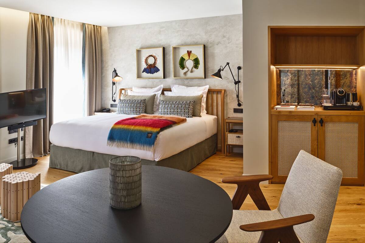 Quarto de hotel moderno com uma cama grande, uma mesa redonda, uma cadeira, uma televisão e uma escrivaninha com uma luminária e livros.
