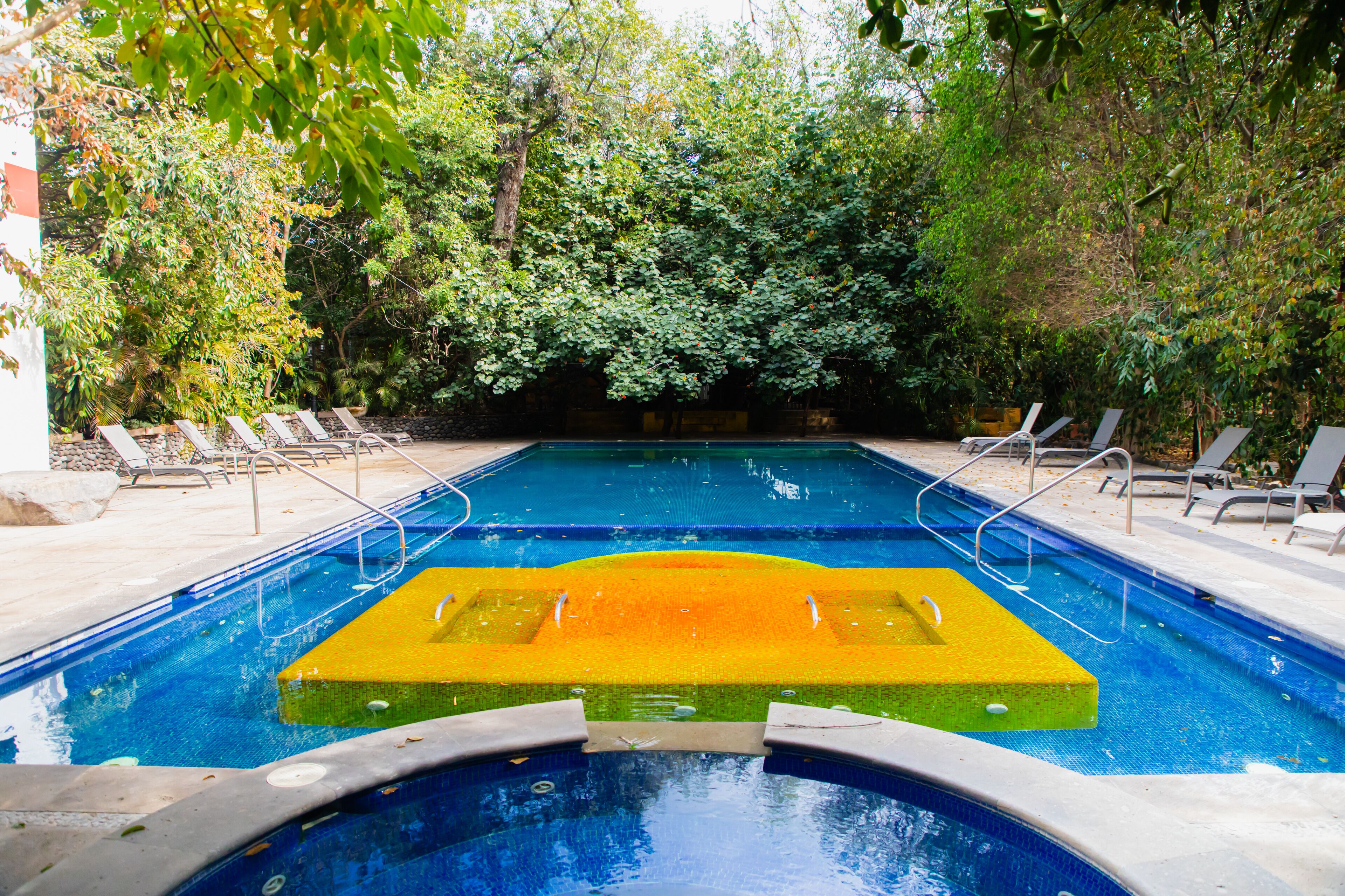 Piscina com plataforma flutuante, rodeada por árvores e espreguiçadeiras.