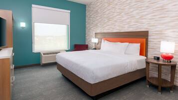 Quarto de hotel com uma cama grande, janela com persianas, televisão fixada na parede, mesa de cabeceira com abajur e uma parede em destaque vermelha.