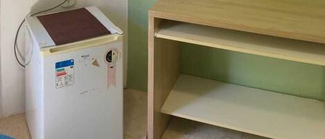 Quarto com televisão na parede, uma pequena prateleira de madeira com uma garrafa, um suporte de madeira para cabides, um eletrodoméstico branco com painel de controle e um banco de madeira.