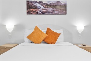 Cama bem arrumada com roupa de cama branca e duas almofadas laranjas, um quadro pendurado na parede de uma cachoeira e duas luzes fixadas na parede, de cada lado da cama.