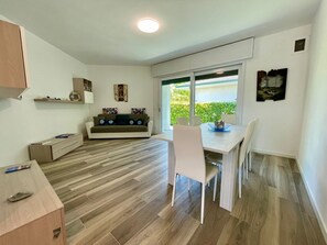 Uma sala de estar moderna com um sofá, mesa de jantar e cadeiras. Há uma janela com vista para o verde lá fora.