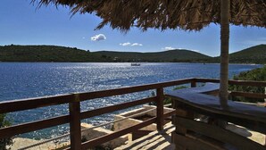 Um deck de madeira com telhado de palha com vista para um corpo de água, com um barco ao longe e colinas ao fundo.