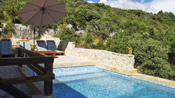Área de piscina com espreguiçadeiras e um guarda-sol, rodeada por um muro de pedra e vegetação exuberante.
