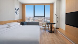 Quarto de hotel moderno com uma cama grande, vista para a cidade, escrivaninha e televisão.