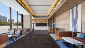 Um saguão de hotel moderno com área de estar central, balcão de recepção e uma grande janela com vista para a cidade.