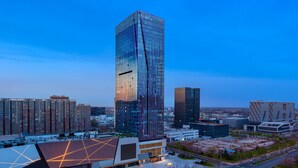 Um edifício moderno de grande altura com fachada de vidro, rodeado por outros edifícios e um parque de estacionamento.