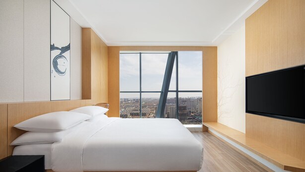 Quarto de hotel moderno com uma cama grande, televisão de tela plana e vista para a cidade através de uma janela ampla.