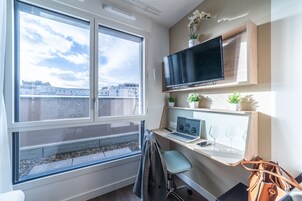 Quarto moderno com uma janela ampla, uma escrivaninha com um laptop, uma televisão de tela plana e uma pequena planta em vaso.