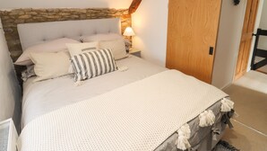 Quarto com parede de pedra, uma cama com vários travesseiros, porta de madeira e mesa de cabeceira com abajur.